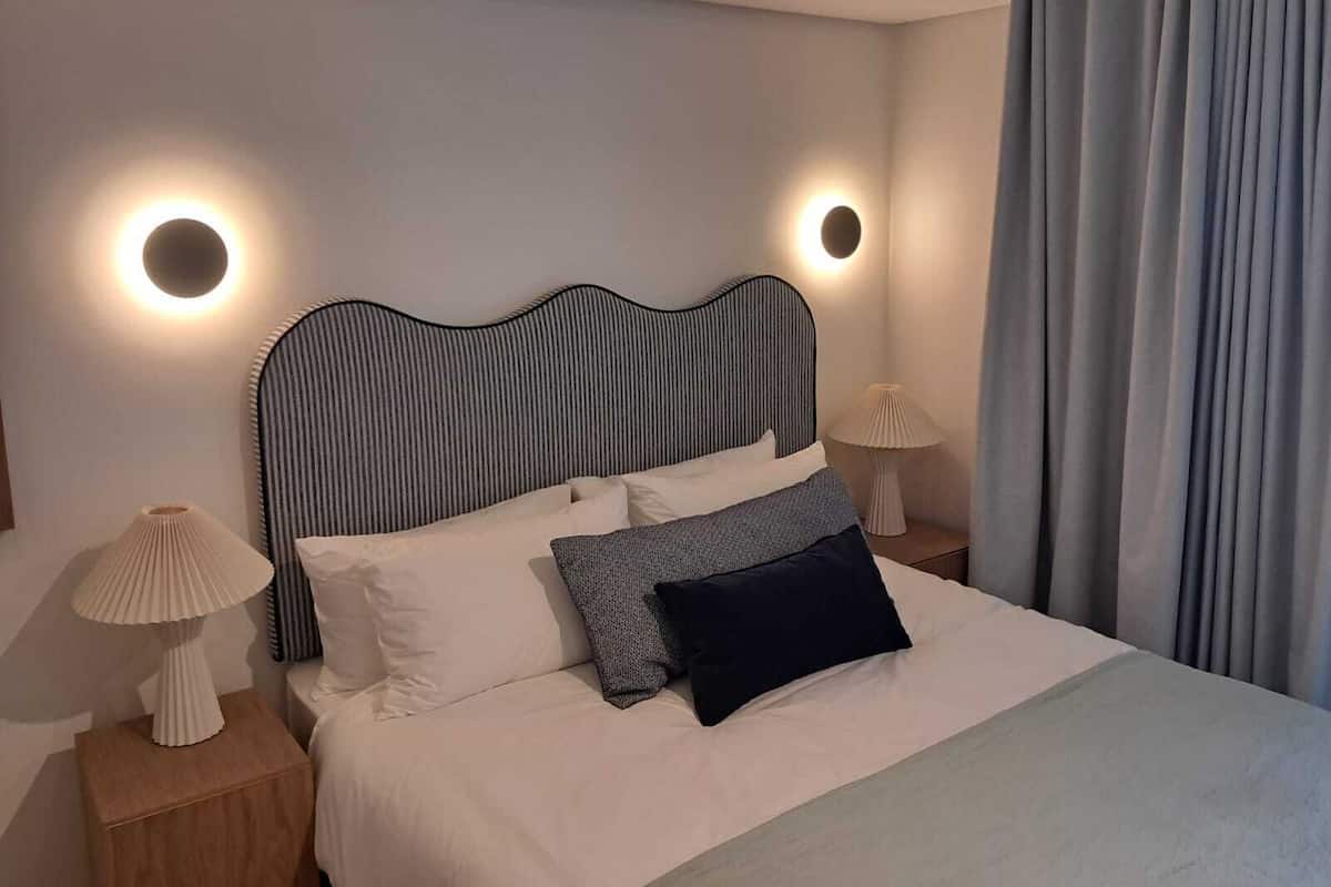 Cama bem arrumada com cabeceira listrada, duas luminárias de cabeceira e uma janela com cortinas.