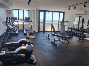 Academia com aparelhos de cardio, pesos livres e vista para o mar.