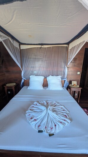 Cama com dossel, duas almofadas, mesa de cabeceira e arranjo decorativo de toalhas.