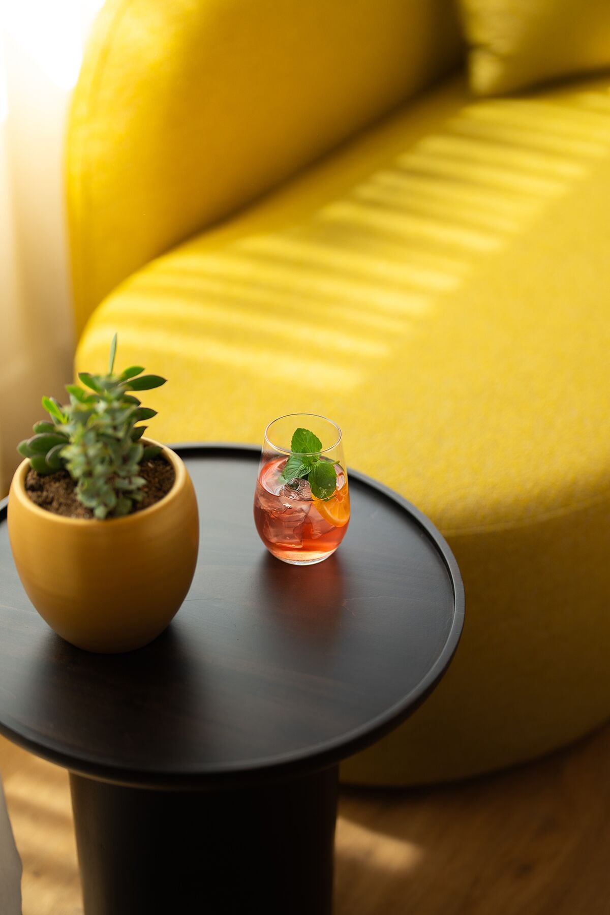 Uma pequena planta em vaso sobre uma mesa redonda, um copo com bebida e guarnição, e um sofá amarelo ao fundo.