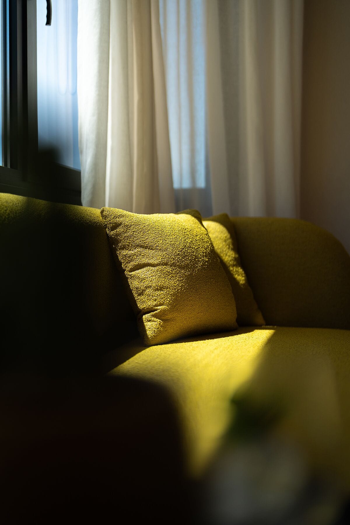 Uma cama com manta e almofadas amarelas, uma janela com cortinas e vista para o céu.