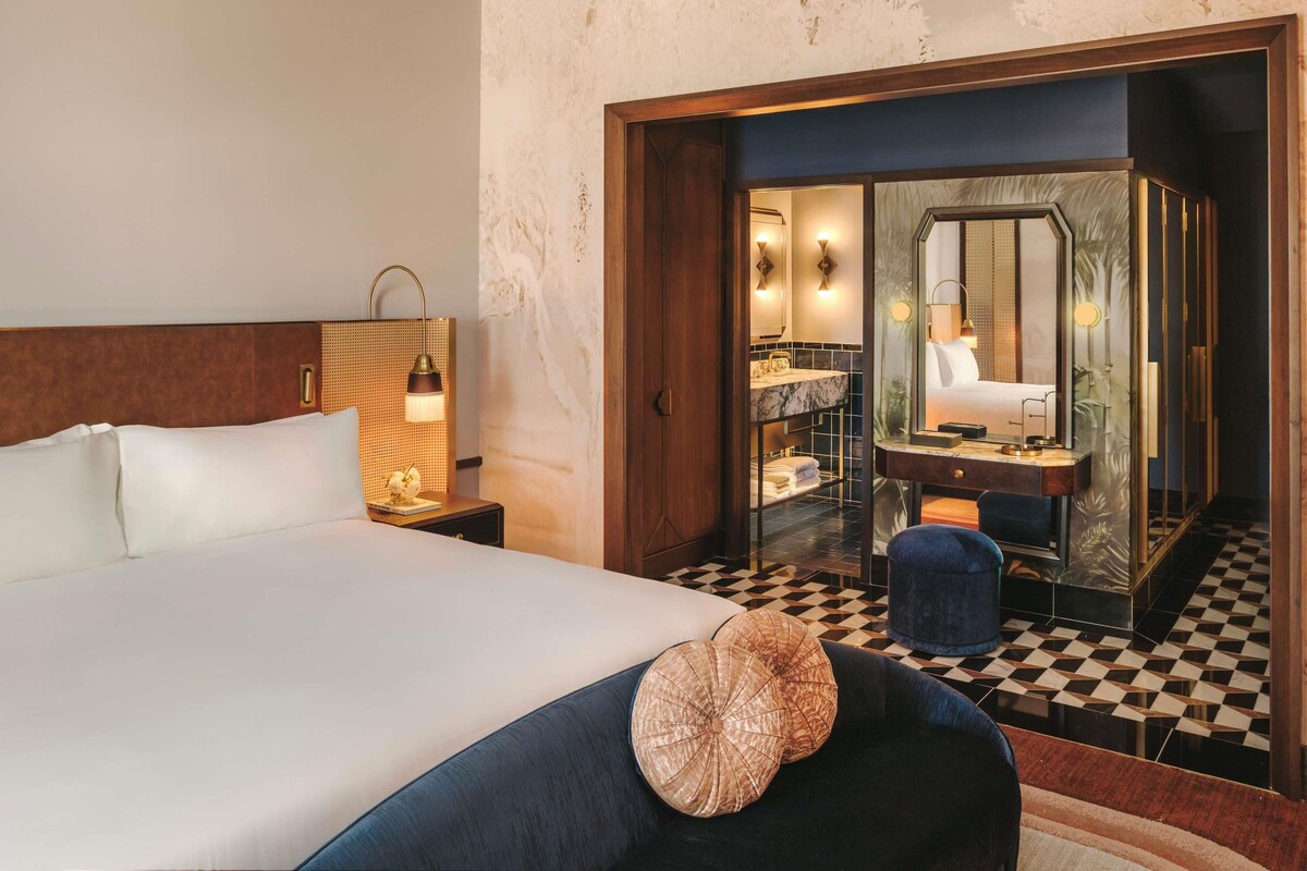Quarto de hotel com uma cama grande, mesa de cabeceira com abajur, banheiro com espelho e pia, e chão xadrez.