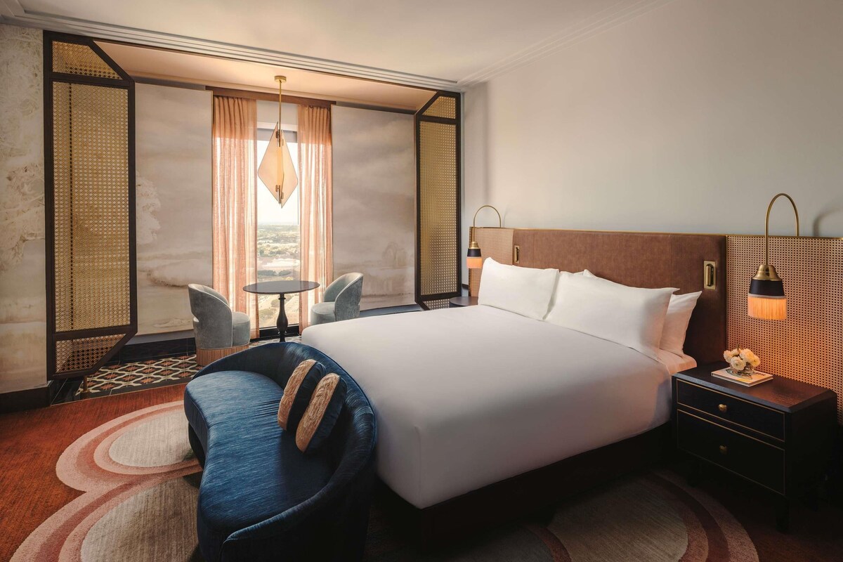 Quarto de hotel com uma cama grande, uma mesinha, duas cadeiras, um criado-mudo e vista pela janela com cortinas.