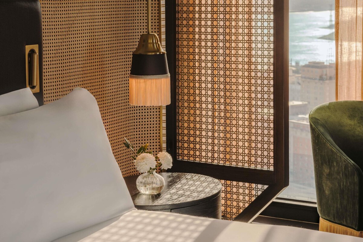 Quarto moderno com cama, mesa redonda com um vaso de flores, poltrona verde e vista para a cidade através de uma janela estampada.