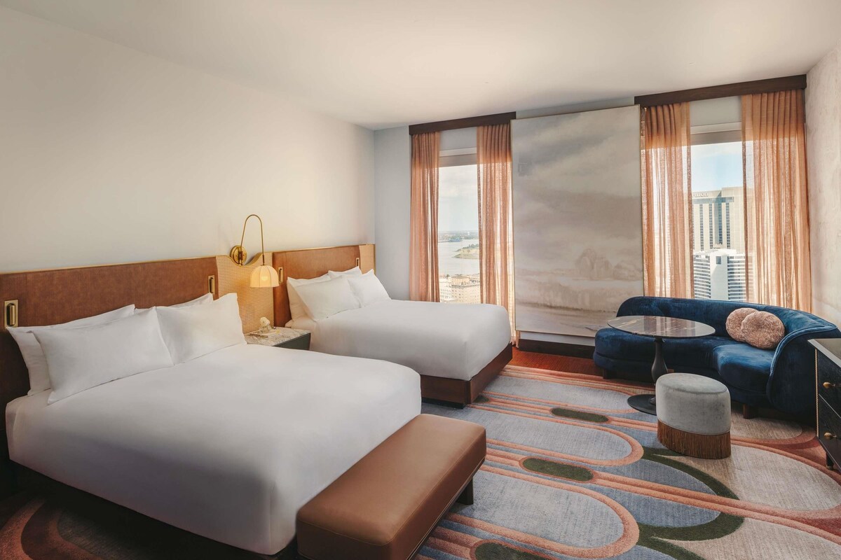 Quarto de hotel com duas camas, um sofá, uma mesa pequena e vista para a cidade.