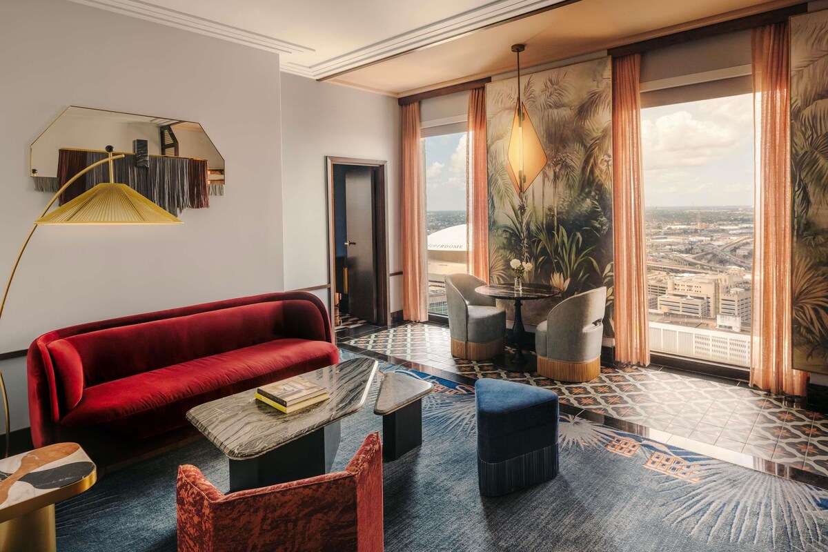 Quarto de hotel moderno com sofá vermelho, mesa de centro redonda e vista para a paisagem urbana através de grandes janelas.