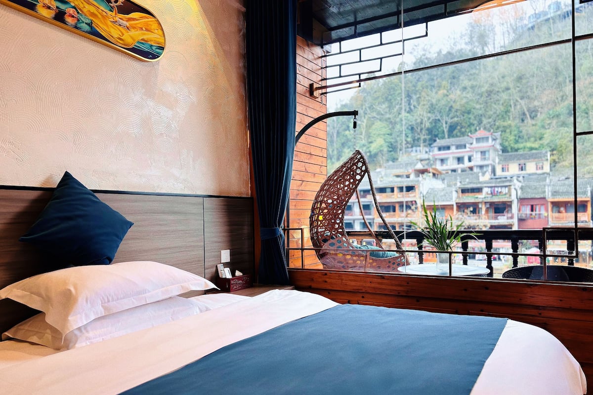 Uma cama com cabeceira, uma janela com vista para prédios, uma cadeira e uma planta.