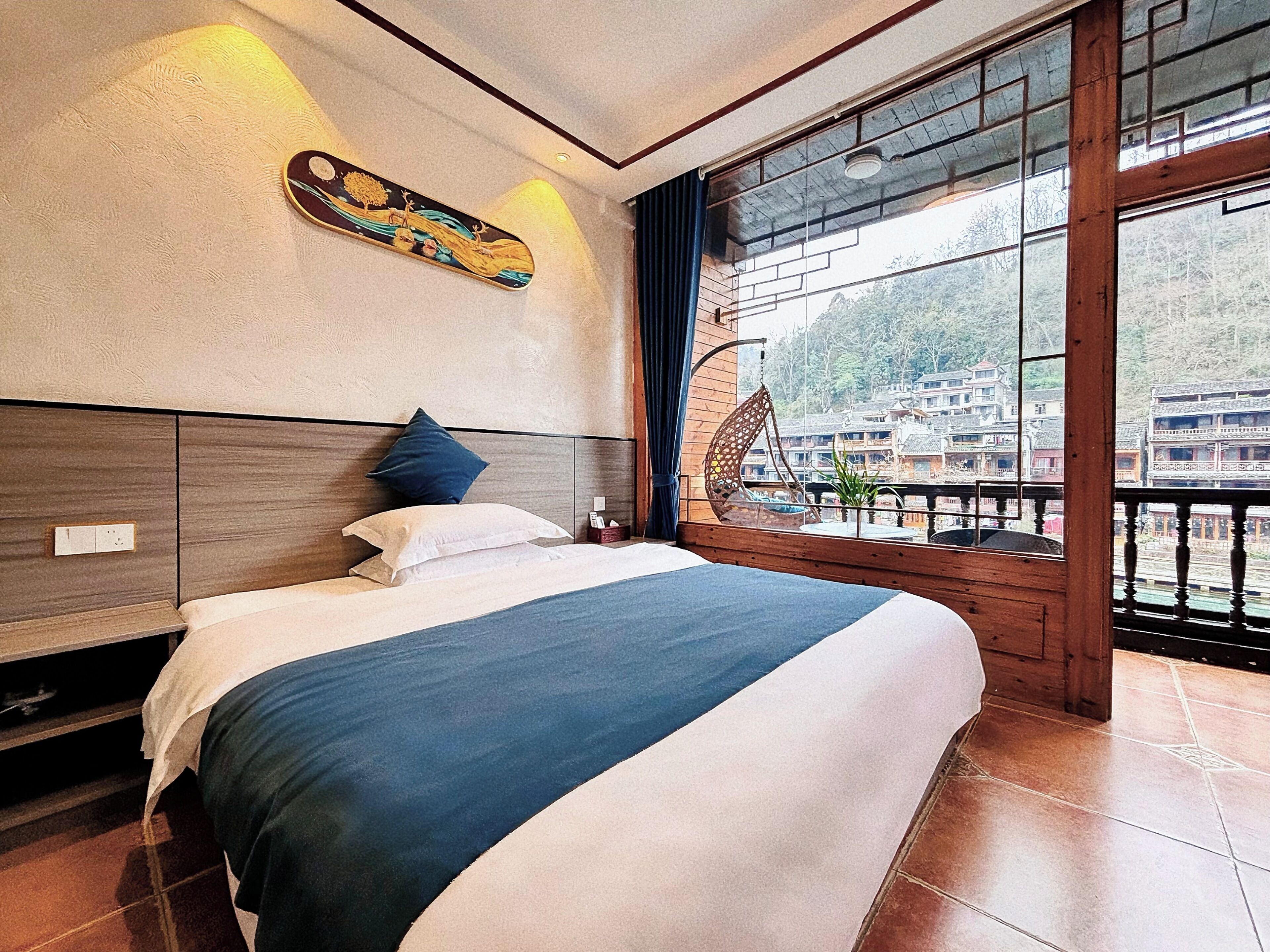 Quarto de hotel com uma cama grande, cabeceira de madeira, uma cadeira suspensa perto da janela e vista para edifícios tradicionais.
