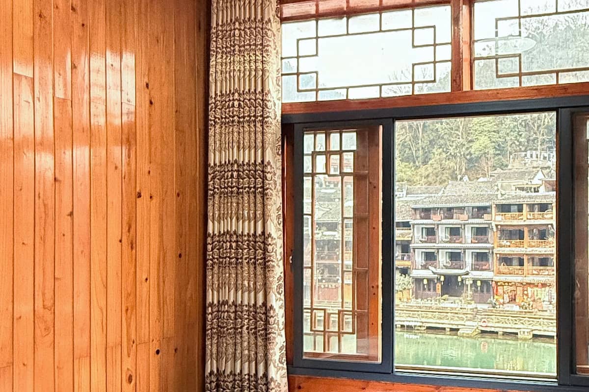 Quarto com paredes de madeira, cama com roupa de cama azul e branca e janela com vista para edifícios tradicionais.