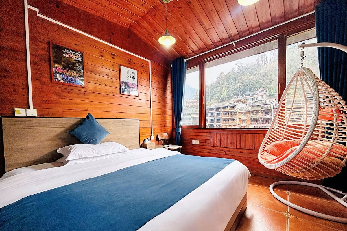 Um quarto de madeira com uma cama, uma cadeira suspensa e uma janela com vista para edifícios.