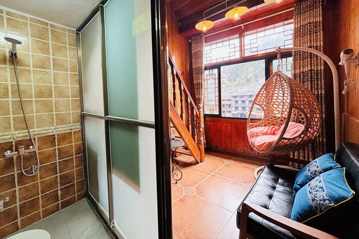 Casa de banho com chuveiro, sanita e lavatório. Escada de madeira leva a uma varanda com uma cadeira suspensa e um sofá.