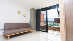 Uma sala de estar moderna com sofá marrom, parede de concreto decorativa e uma grande porta de vidro deslizante que dá para uma varanda com parede de tijolos e vista para as montanhas.