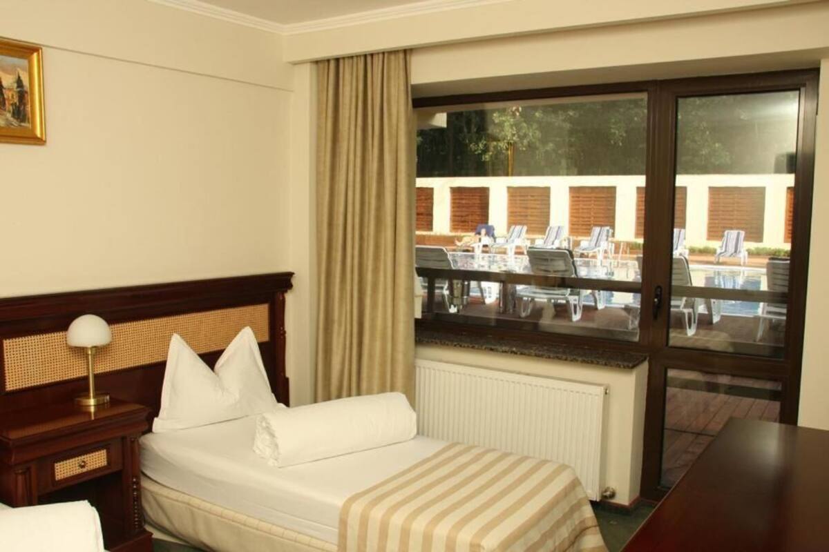 Quarto de hotel com cama, mesa de cabeceira, abajur e janela com vista para uma área externa com mesas e cadeiras.