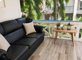 Um sofá de couro preto com uma almofada, uma pequena mesa de madeira com um candeeiro e um corrimão branco com vegetação ao fundo.