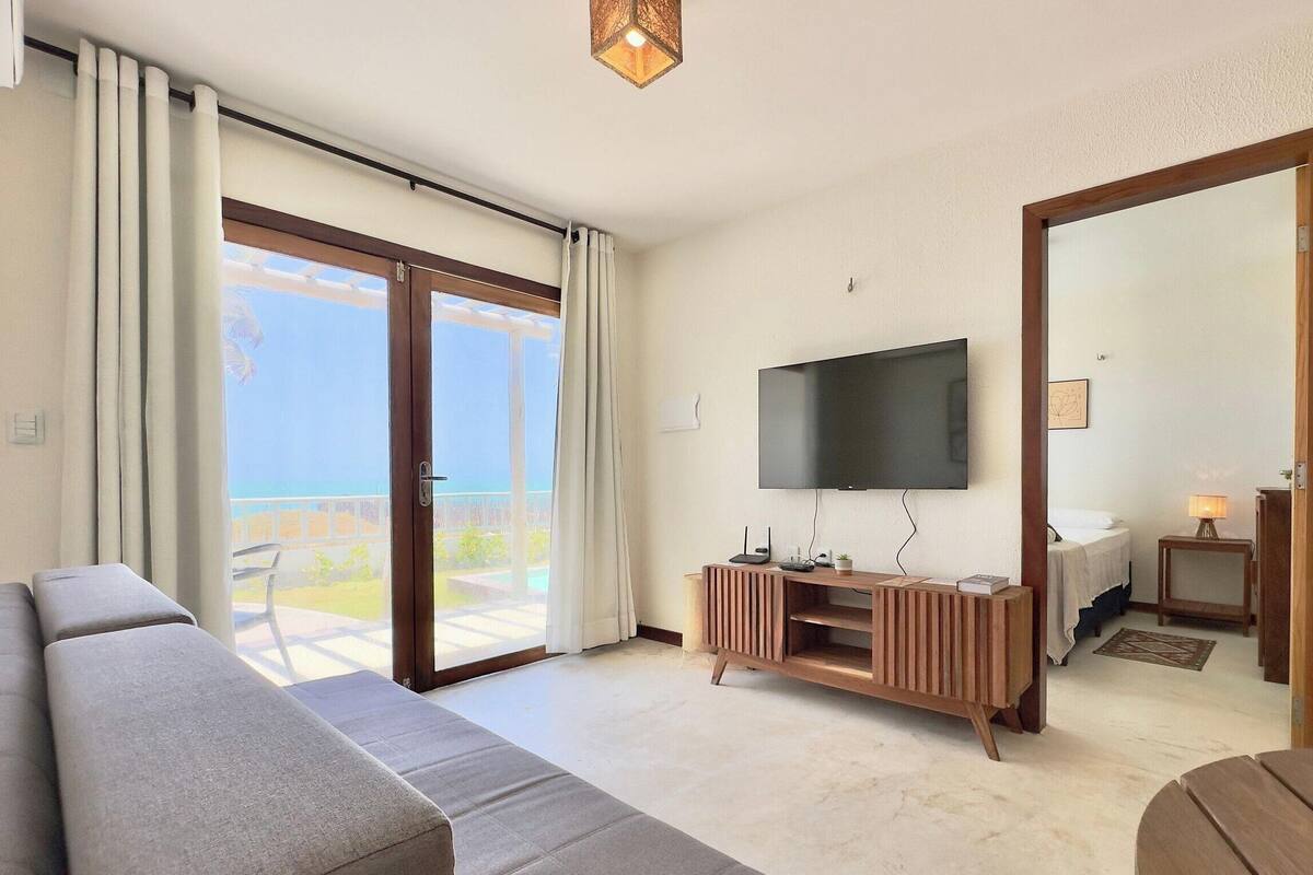 Quarto de hotel com sofá, televisão, mesa consola de madeira e varanda com vista.