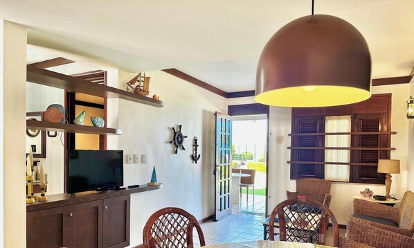 Sala de estar com televisão, cadeiras de vime, mesa de jantar, luminária pendente grande e vista para o exterior.