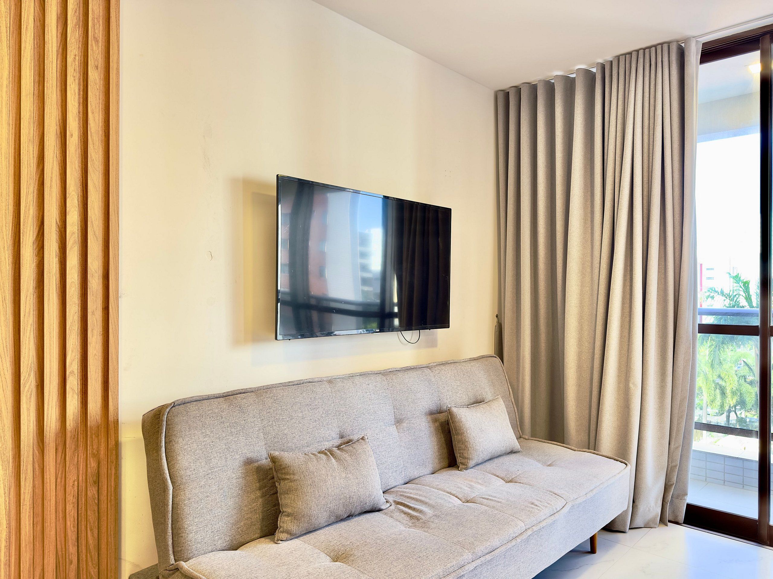 Sala de estar moderna com sofá, TV fixada na parede e uma grande janela com cortinas.