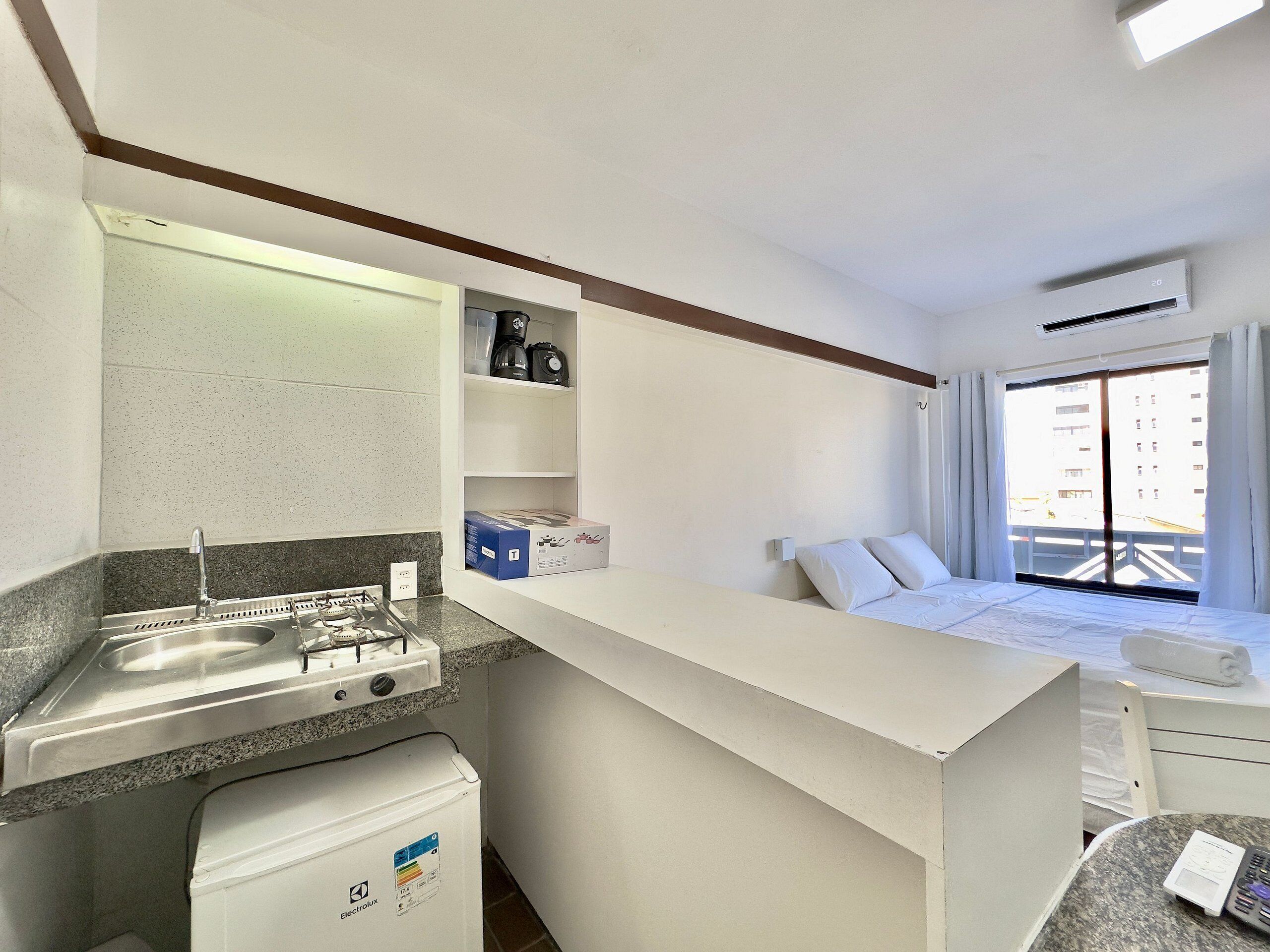Quarto de hotel pequeno com cozinha compacta, uma cama com almofadas, janela com cortinas e vista para um edifício.