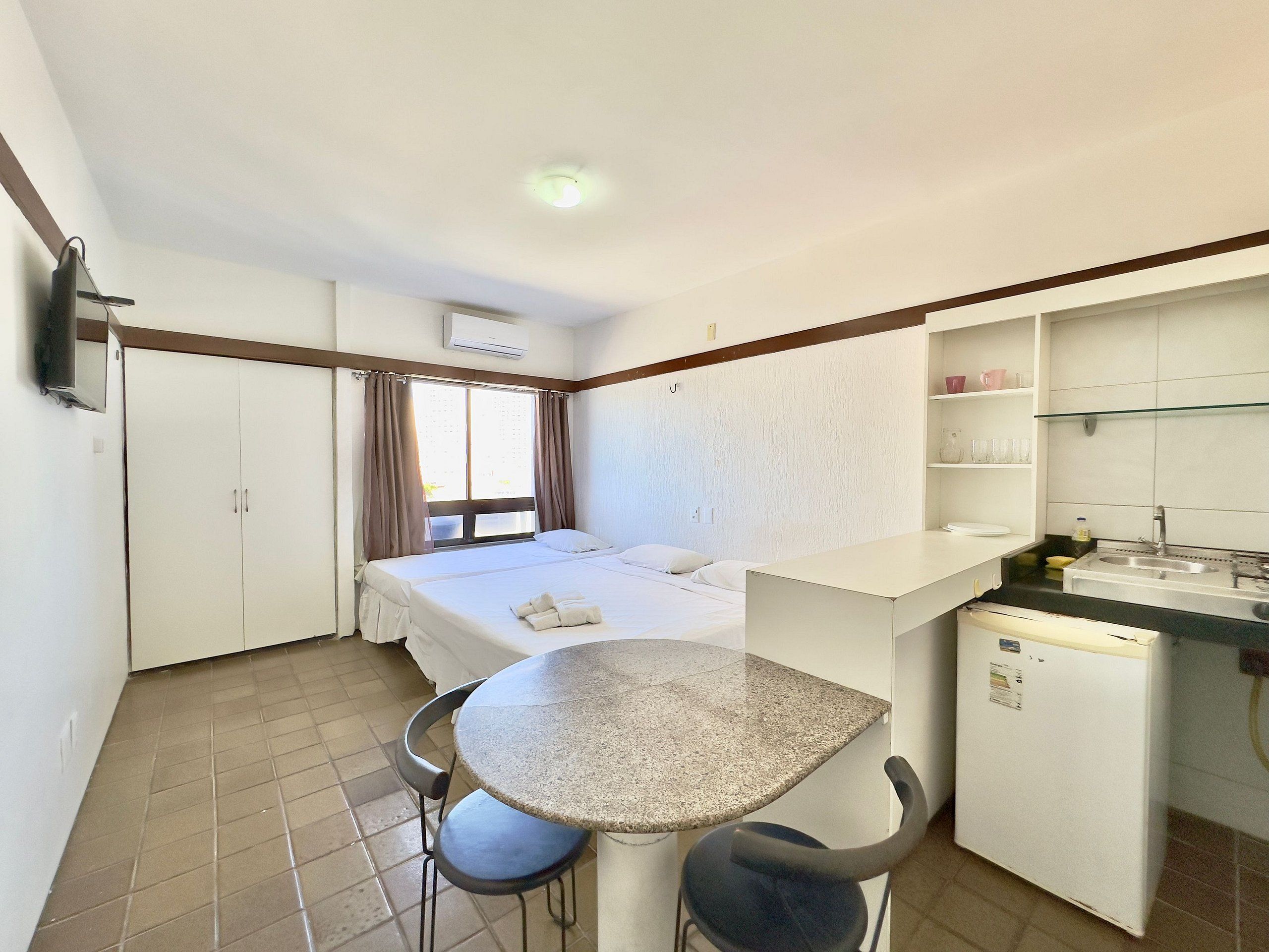 Quarto de hotel compacto com cama, pequeno espaço de cozinha, área de refeições com mesa e cadeiras, e janela com cortinas.
