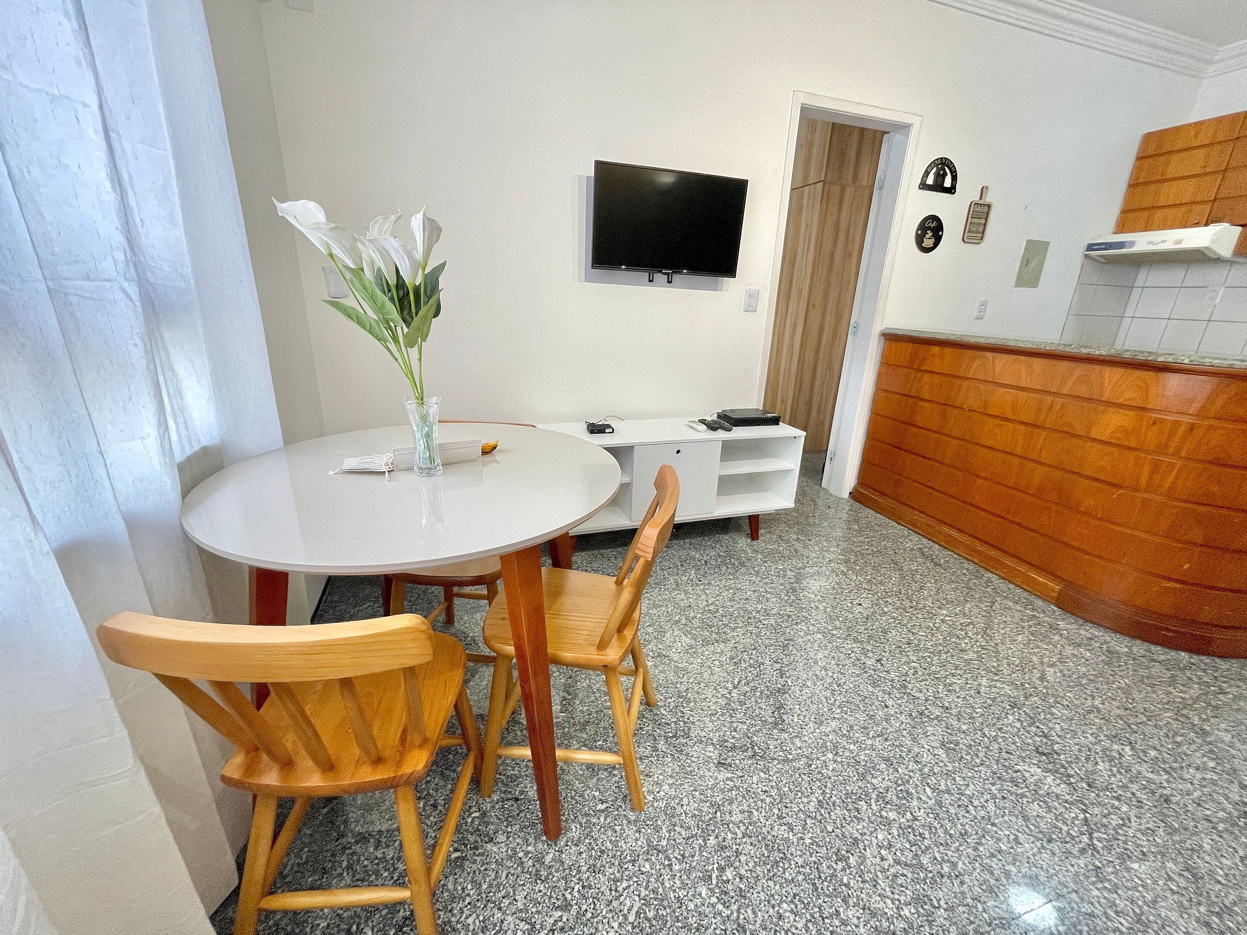 Uma área de refeições com uma mesa redonda, duas cadeiras de madeira, um vaso com flores, uma televisão de tela plana na parede e um balcão de madeira.