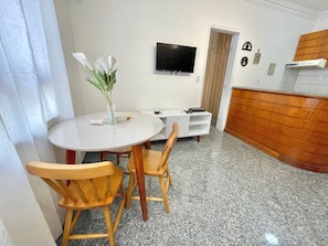 Uma área de refeições com uma mesa redonda, duas cadeiras de madeira, um vaso com flores, uma televisão de tela plana na parede e um balcão de madeira.