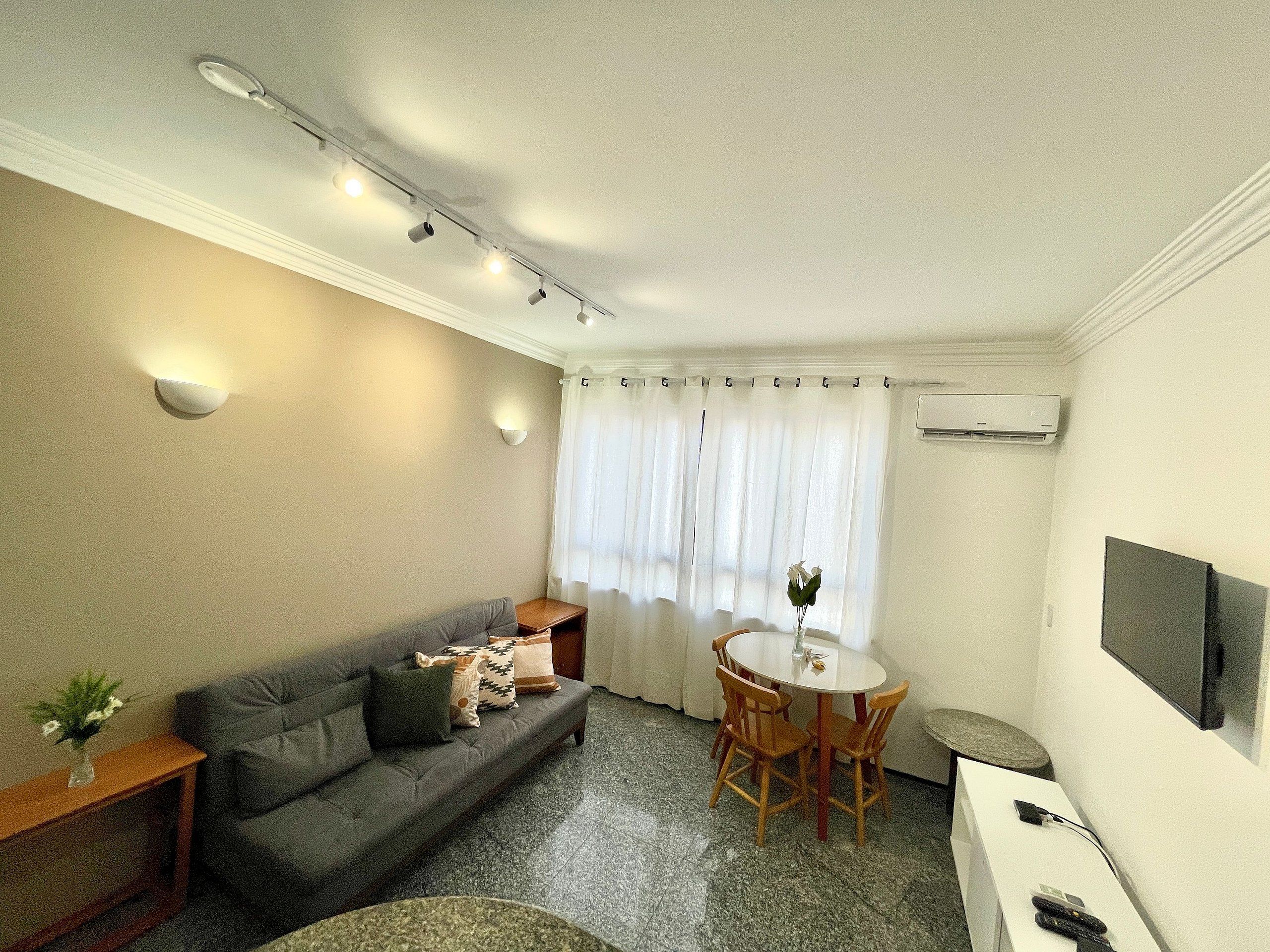Uma sala de estar com sofá, mesa pequena, mesa de jantar com cadeiras, televisão e ar condicionado.