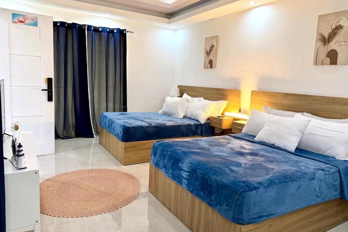 Quarto de hotel com duas camas, uma escrivaninha, uma luminária e uma parede com obras de arte.