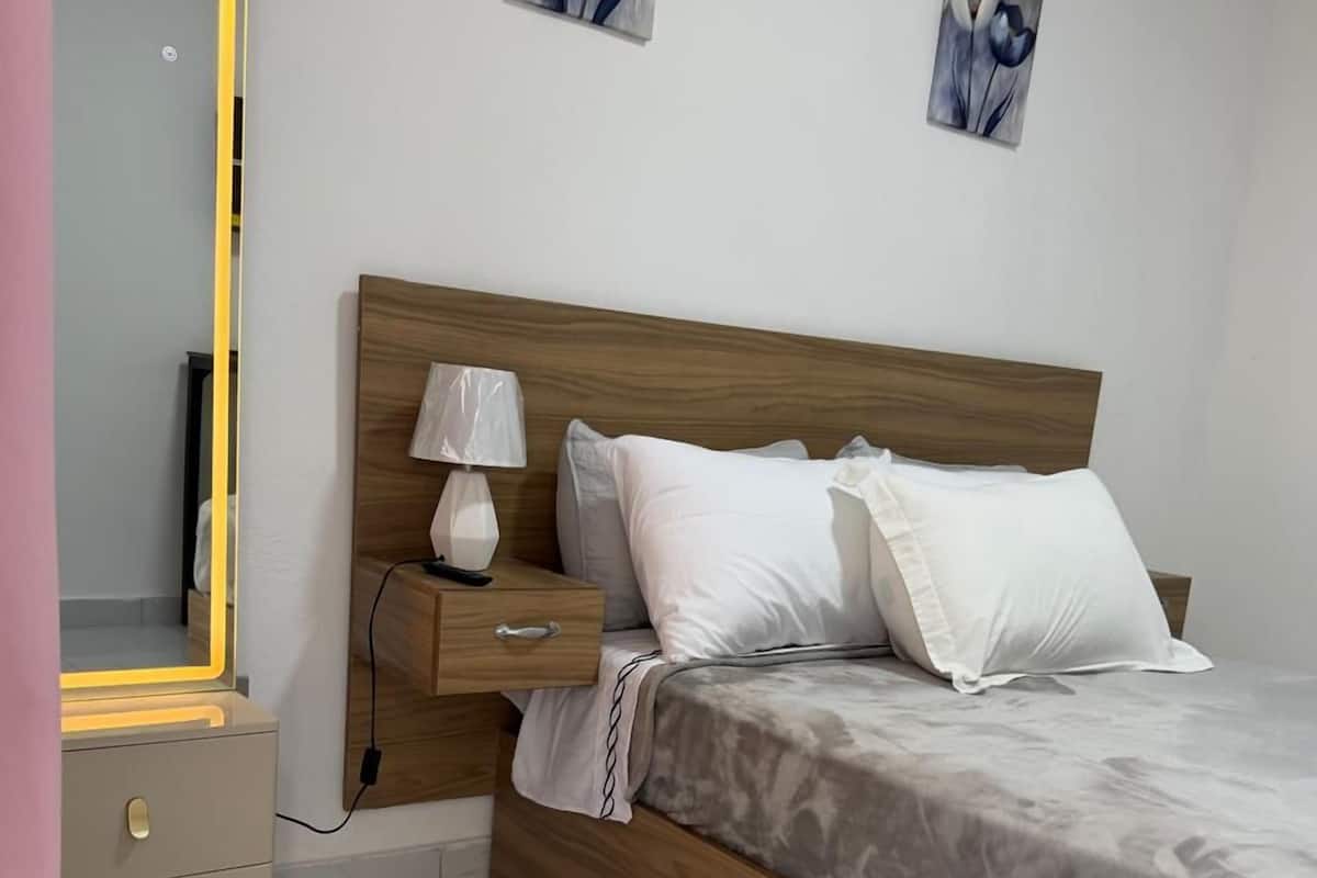 Quarto moderno com cabeceira de madeira, mesa de cabeceira com abajur, espelho e duas pinturas nas paredes.