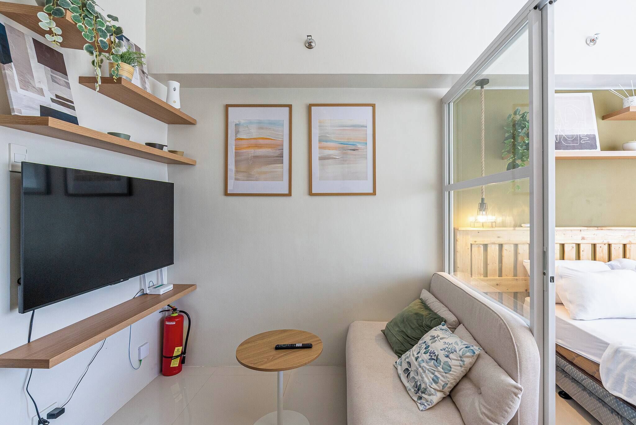 Uma sala de estar moderna com televisão de tela plana, prateleiras de madeira, duas obras de arte emolduradas, uma mesa redonda pequena, um sofá com almofadas e uma porta de vidro deslizante que dá para uma varanda.