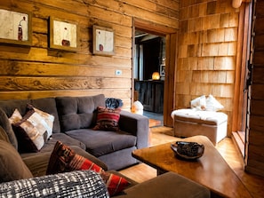 Interior acolhedor de uma cabana de madeira, com sofá, apoio para os pés e uma mesa com um recipiente de pinhas.