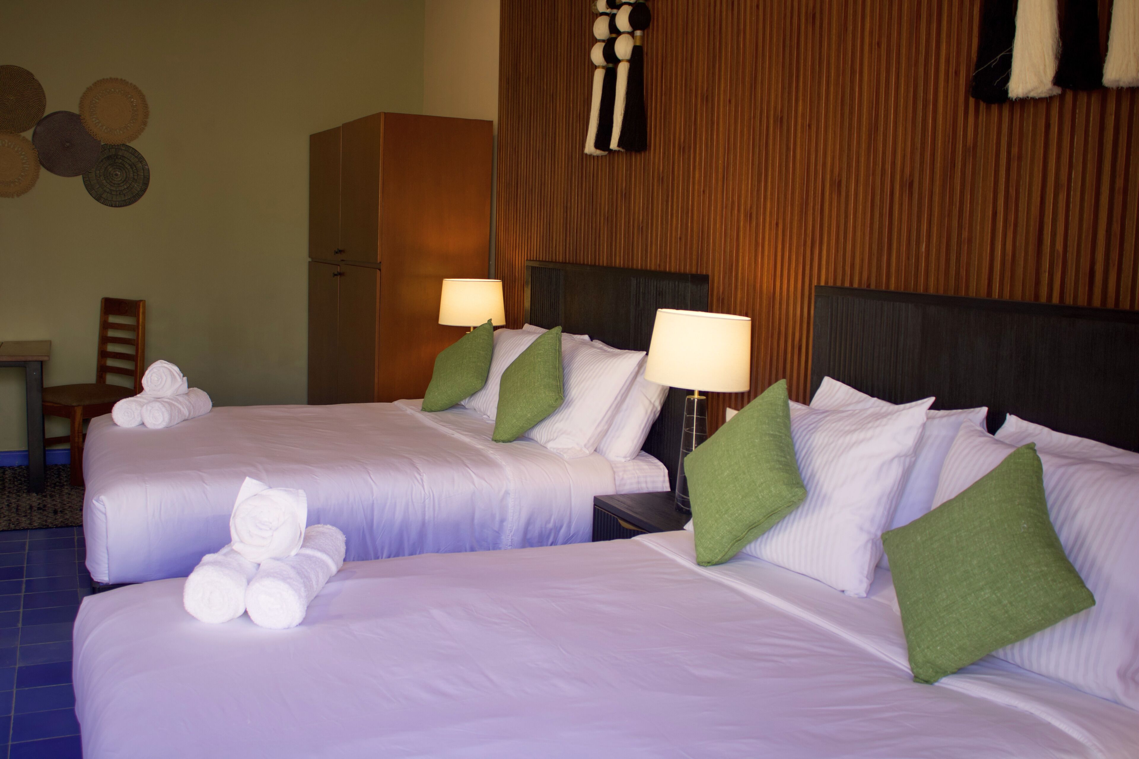 Quarto de hotel com duas camas, cada uma com roupa de cama branca e almofadas verdes, cabeceira de madeira e uma parede com design de painéis verticais de madeira.