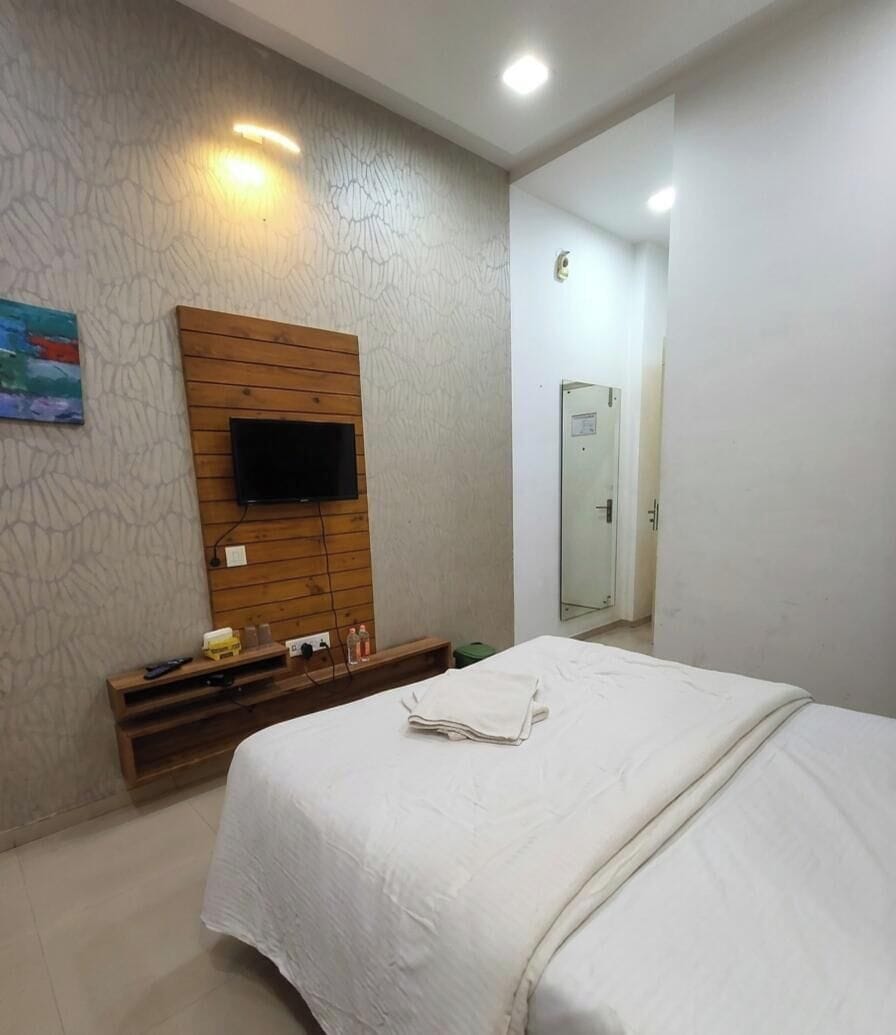 Quarto com cama, televisão presa a um painel de madeira, prateleira pequena com artigos de higiene e porta para um banheiro.