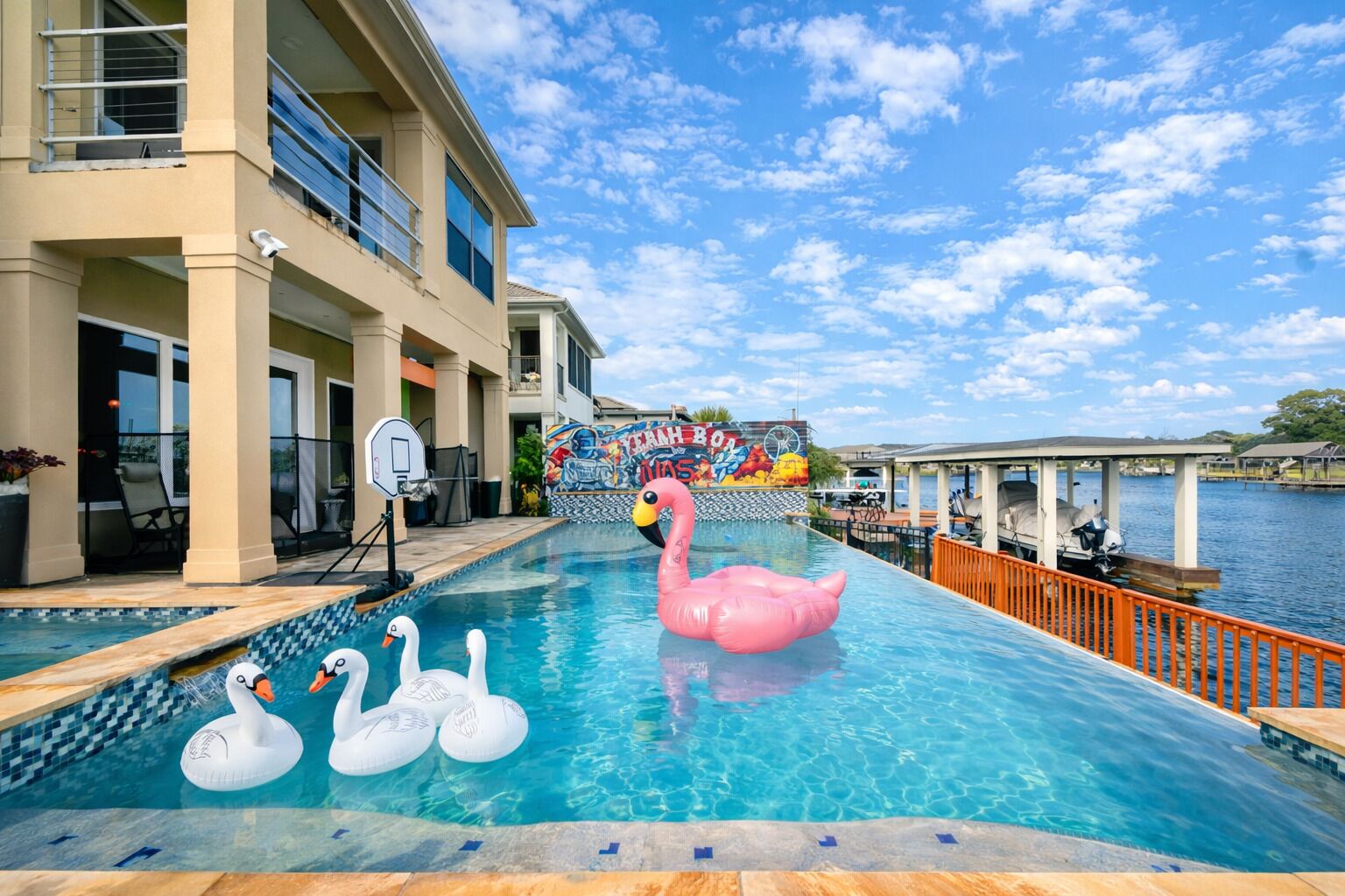 Piscina com boias em formato de cisne e flamingo, um aro de basquete e um mural no edifício.