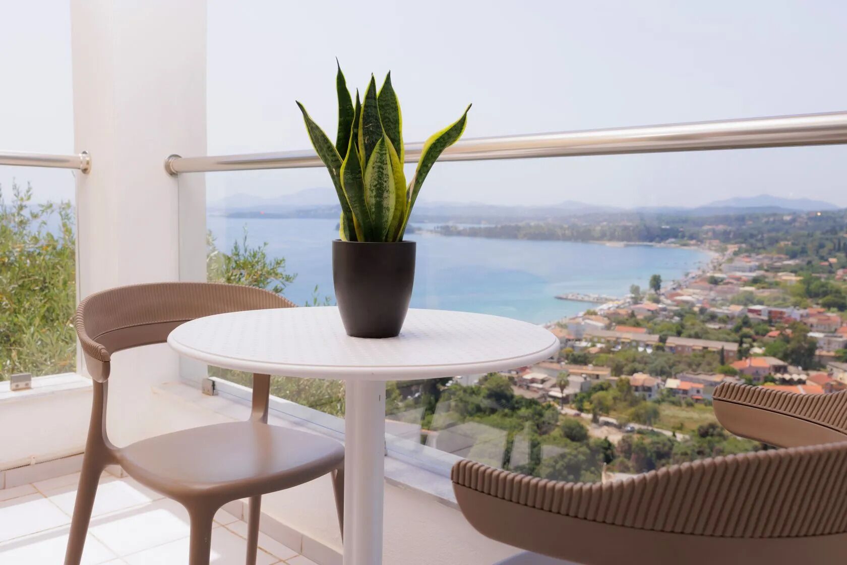 Varanda com vaso de planta, mesa redonda e duas cadeiras com vista para uma cidade costeira.
