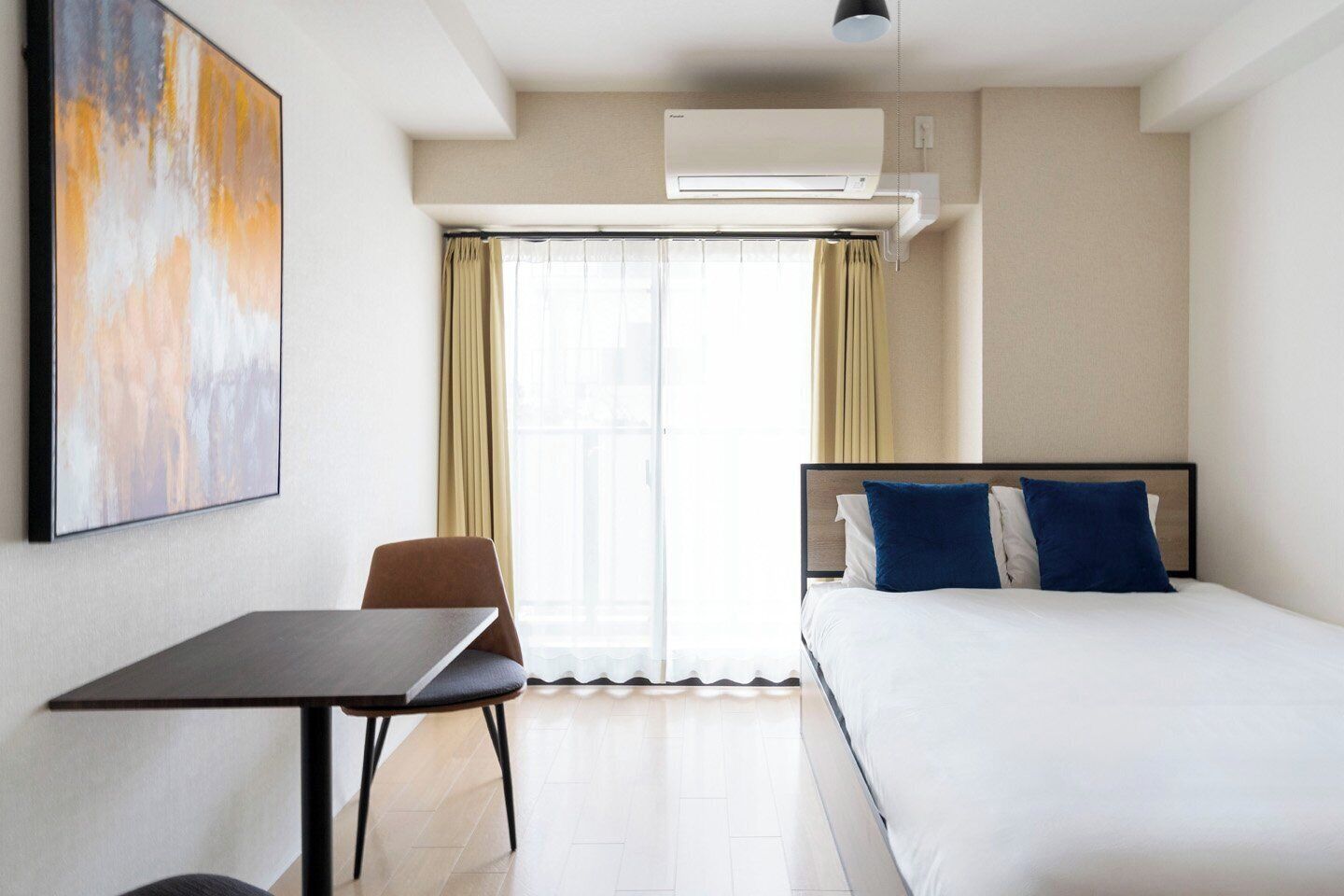 Quarto de hotel com cama, escrivaninha, cadeira, janela com cortinas e ar condicionado.