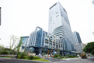 Um edifício moderno com muitos andares e janelas de vidro refletivo, uma entrada destacada e área ao redor bem cuidada, com vegetação e uma estrada visível.