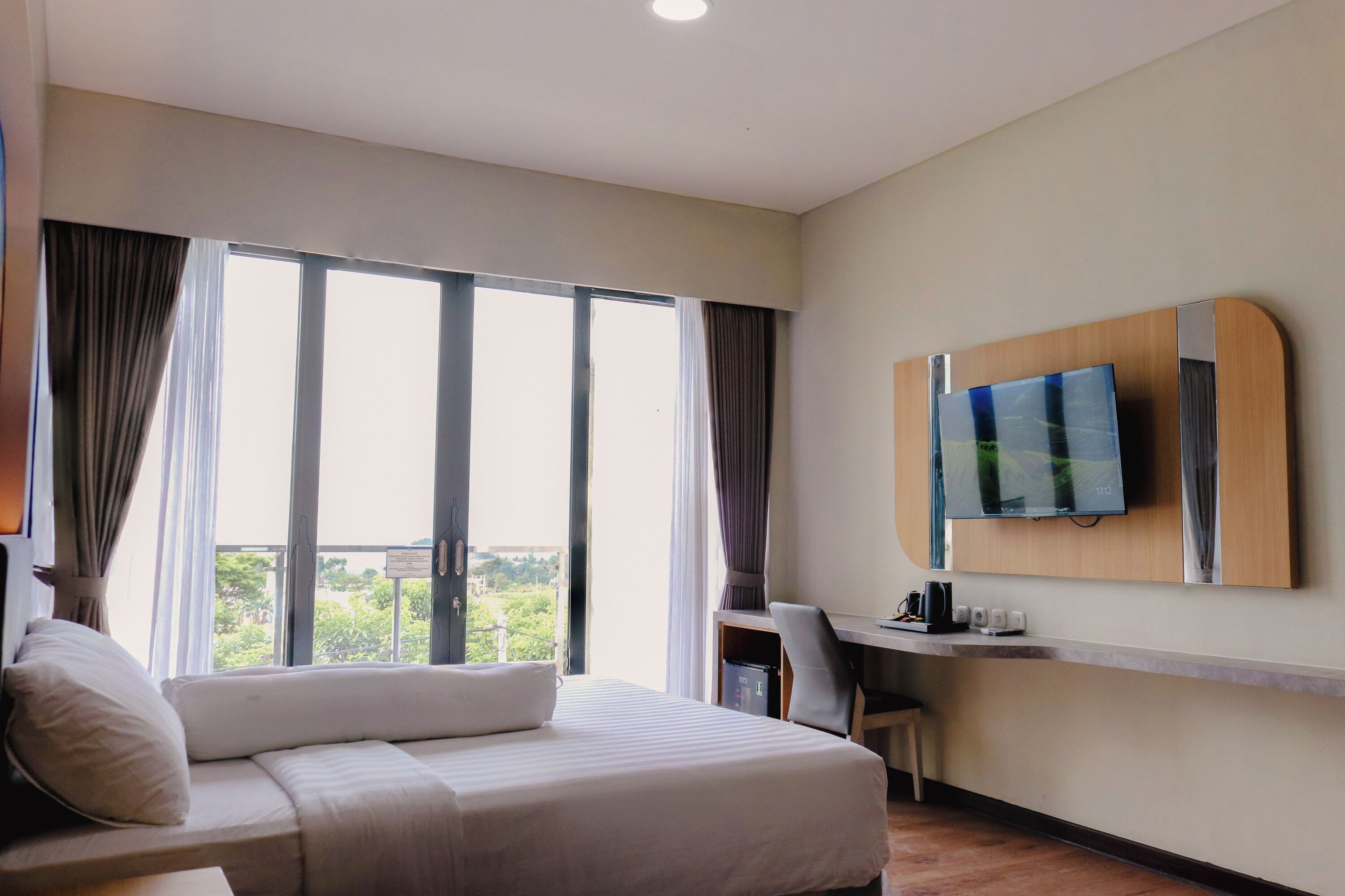 Quarto de hotel com cama, escrivaninha, cadeira, televisão e amplas janelas.