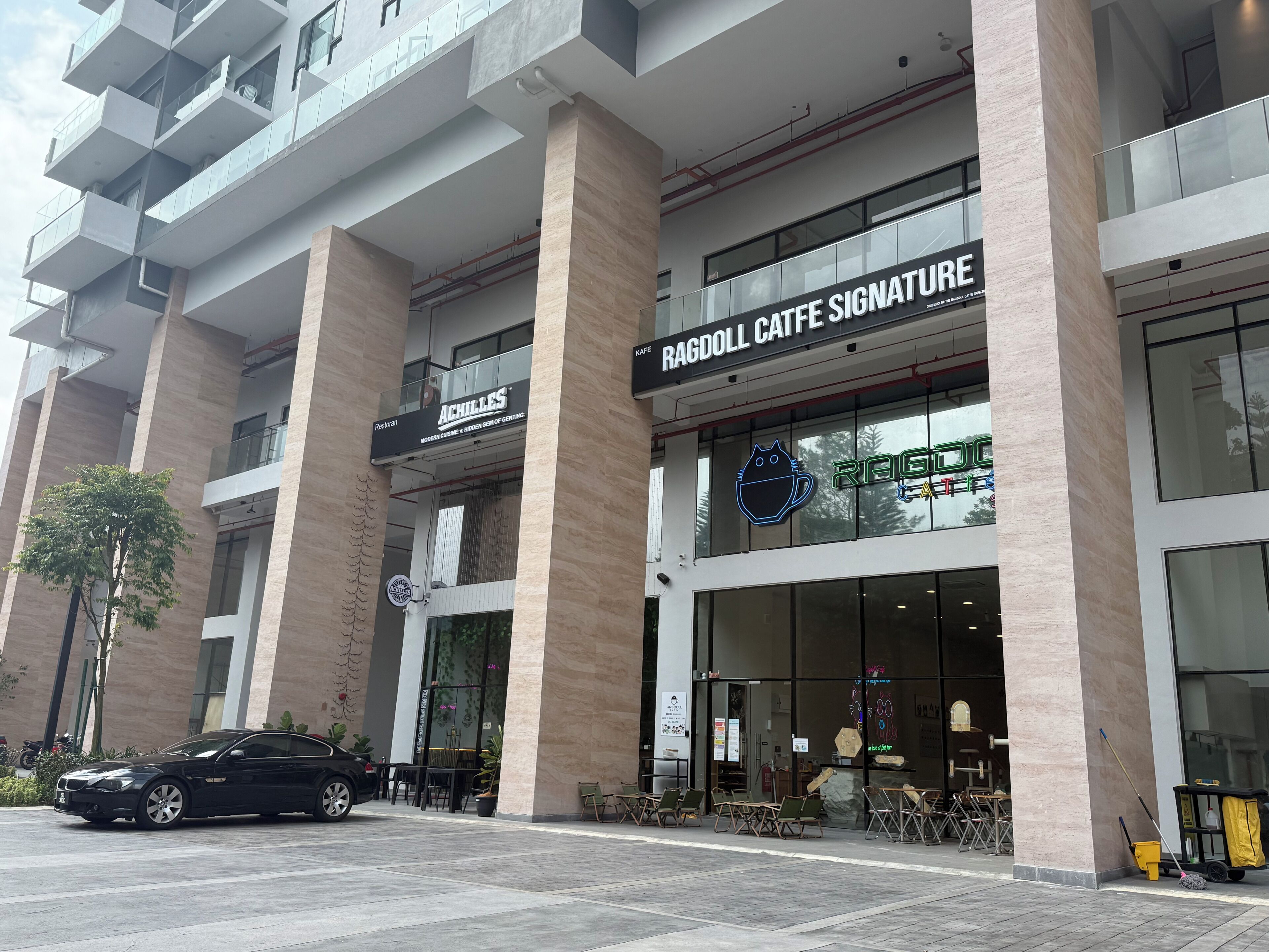 Um edifício moderno com um restaurante chamado 'ACHILES' e uma cafeteria chamada 'RAGDOLL CAFFE SIGNATURE'.