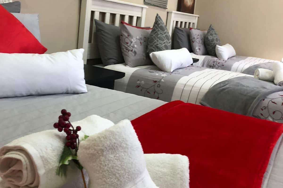 Quarto de hotel com duas camas, cada uma com um cobertor vermelho e branco, e um edredom cinza e branco estampado. Há várias almofadas em cores e padrões diversos.