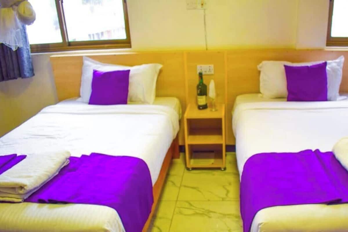 Quarto de hotel com duas camas, cada uma com colcha roxa, uma mesinha de cabeceira de madeira entre elas e uma garrafa de vinho sobre a mesinha.