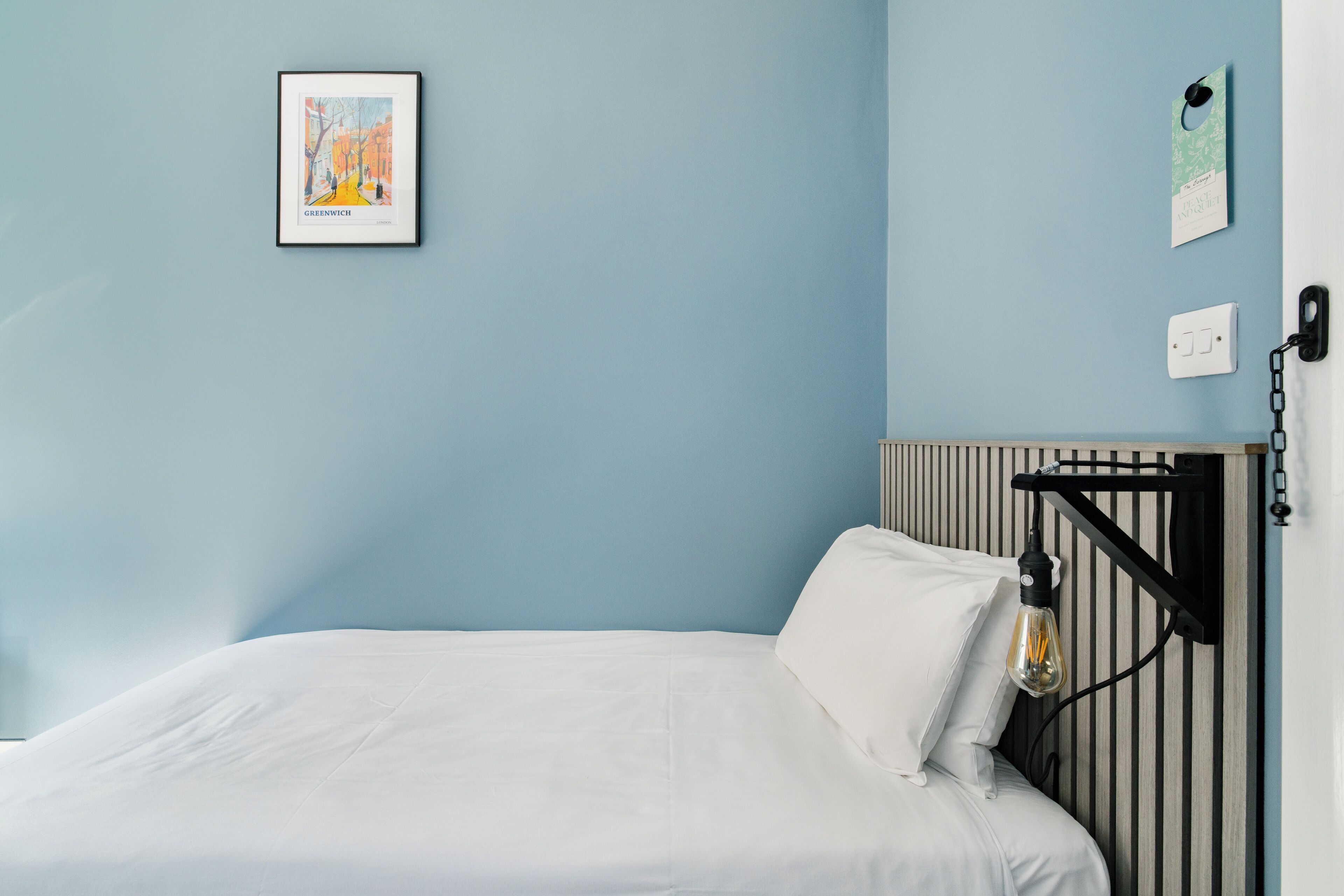 Uma cama com roupa de cama branca, um quadro na parede e uma luminária fixada na parede.