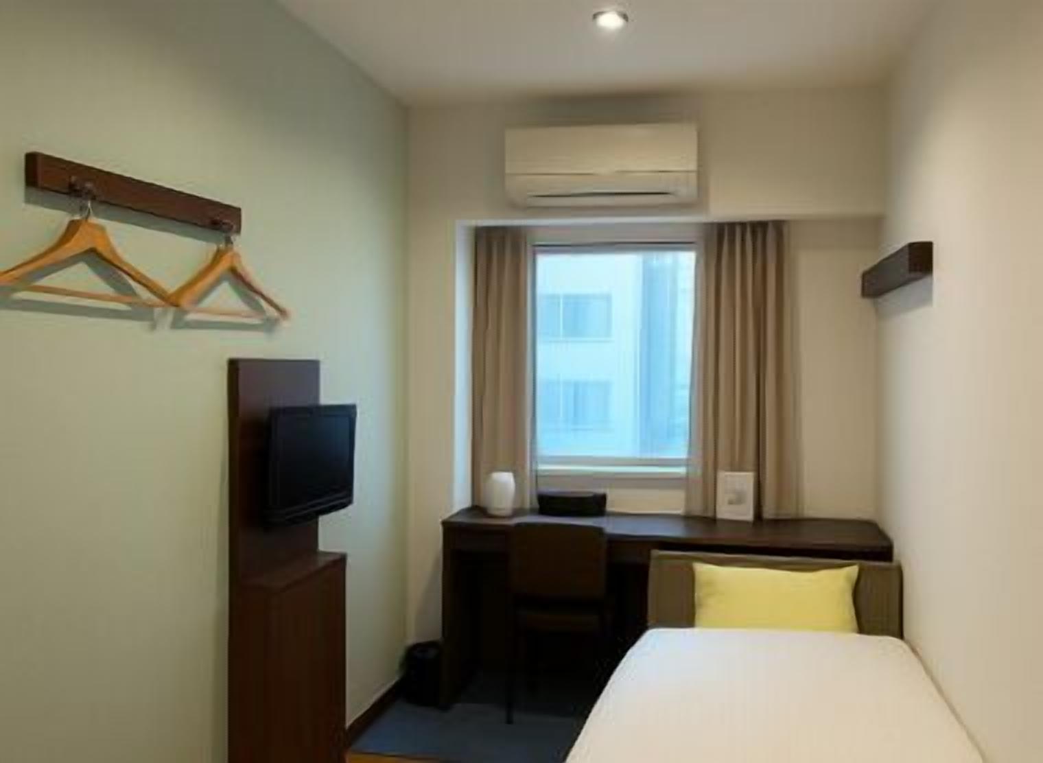 Quarto de hotel com cama, escrivaninha, televisão, janela com cortinas e aparelho de ar condicionado.