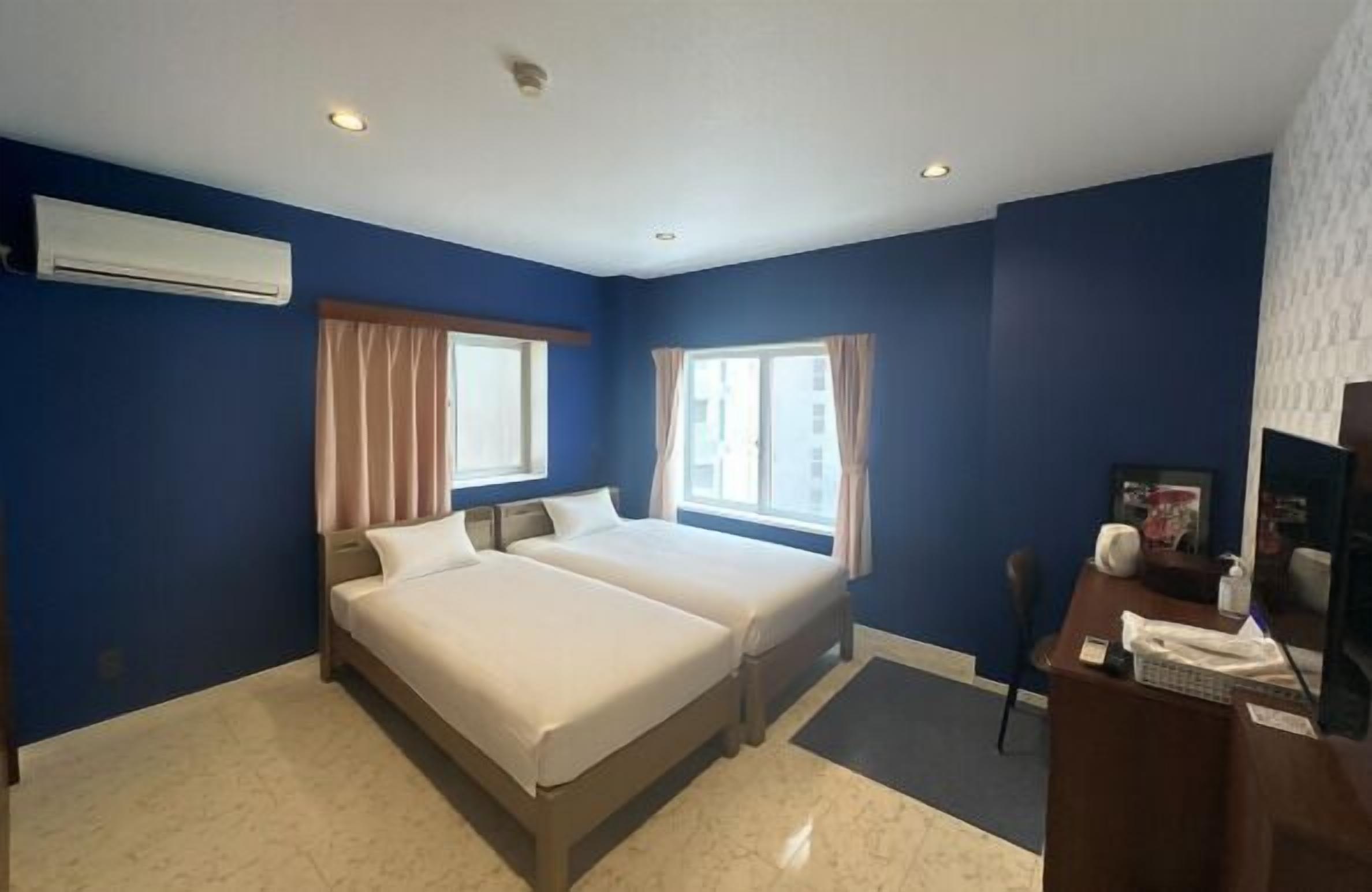 Quarto de hotel com duas camas, uma mesa, uma cadeira, ar condicionado e janela com cortinas.