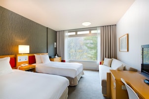 Quarto de hotel com duas camas, uma escrivaninha, televisão e uma janela grande com cortinas.