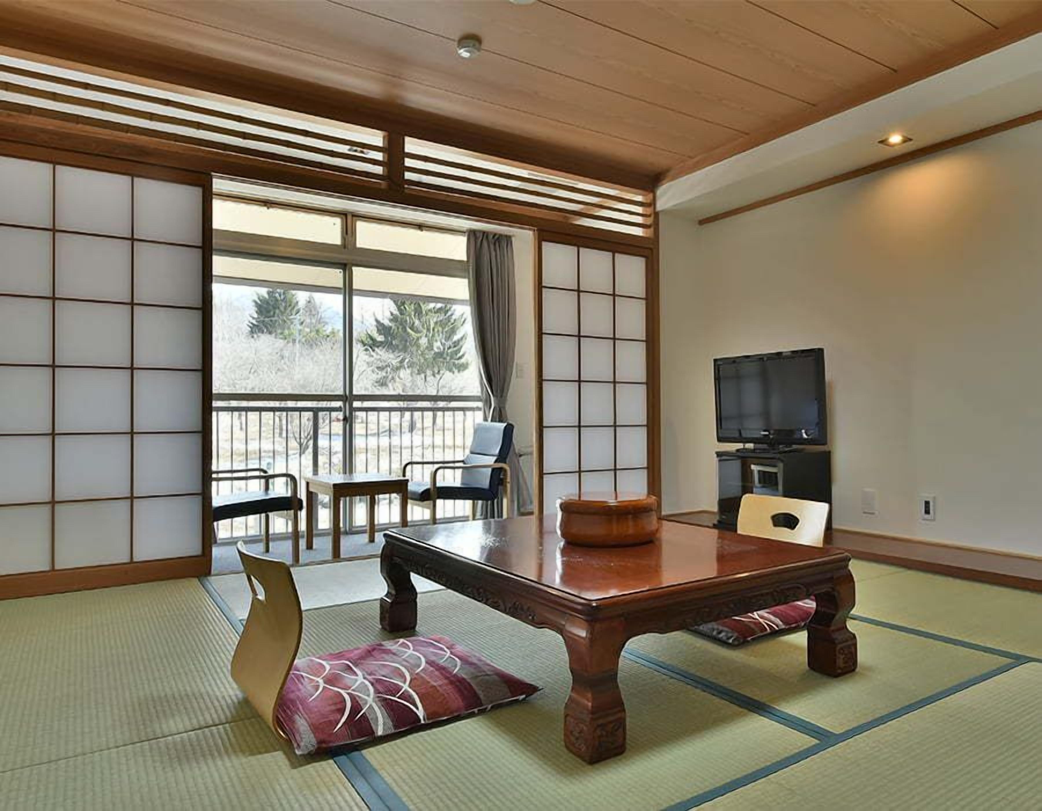 Um quarto em estilo japonês tradicional, com tatames, uma mesa de madeira baixa, televisão e vista para o exterior.