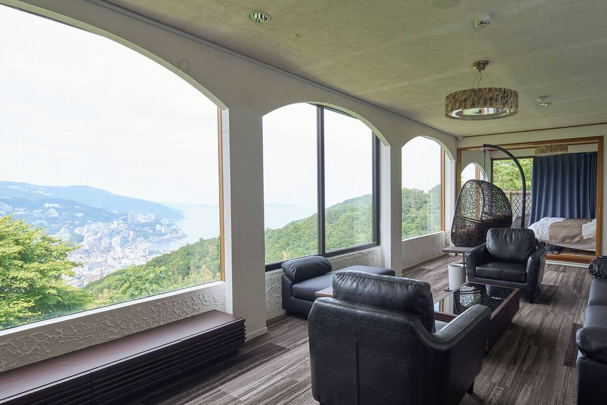 Quarto espaçoso com uma grande janela que oferece uma vista panorâmica de uma cidade e montanhas. Há uma área de estar com sofá e cadeiras, uma cadeira suspensa e uma cama com vista.