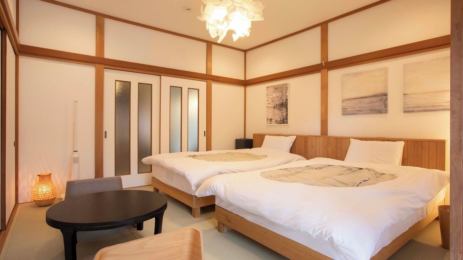 Quarto de hotel com duas camas, uma mesa pequena, uma cadeira, um abajur e obras de arte nas paredes.