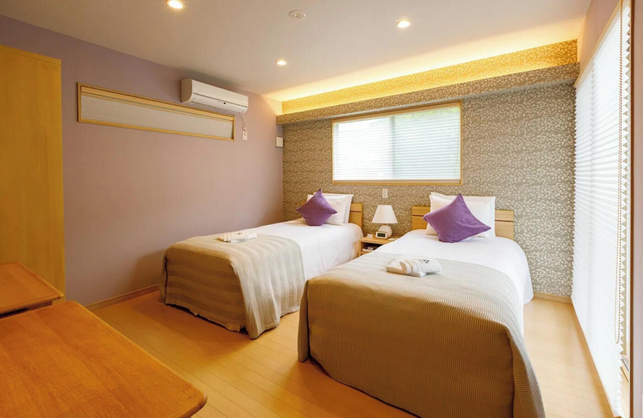 Quarto de hotel com duas camas, uma mesa de madeira, ar condicionado e janela com persianas.