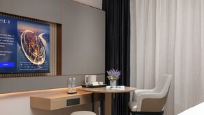 Quarto de hotel moderno com um grande ecrã digital, uma mesa redonda com uma cadeira, uma mesa de cabeceira com uma lâmpada e uma janela com cortinas leves.