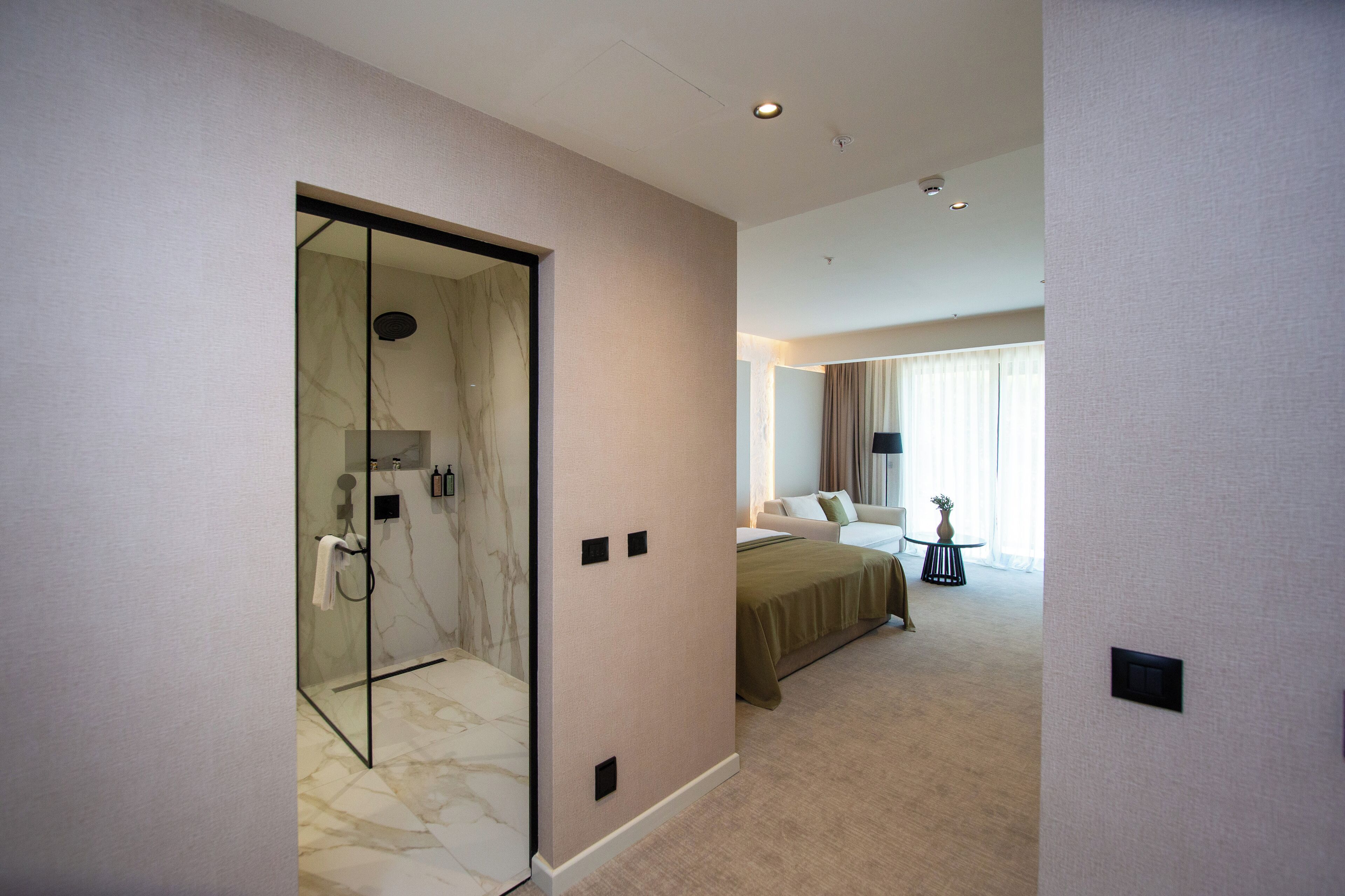 Quarto de hotel com chuveiro em gabinete de vidro, cama com colcha verde, mesa de cabeceira com um vaso e uma janela grande com cortinas.